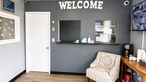 Uma área de recepção com um letreiro 'WELCOME', um balcão de atendimento com espelho, uma cadeira, uma máquina de café e um mural de avisos.
