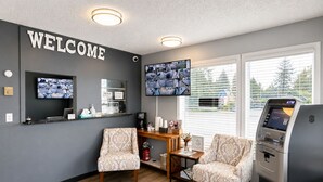 Uma área de recepção com um letreiro 'WELCOME', um balcão de atendimento, duas cadeiras estampadas, uma máquina de café, uma TV exibindo diversas câmeras e um caixa eletrônico.