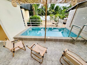 Piscina com deck azulejado, rodeada por palmeiras e um cercado branco. Há três cadeiras dobráveis bege e uma escada de metal para piscina.