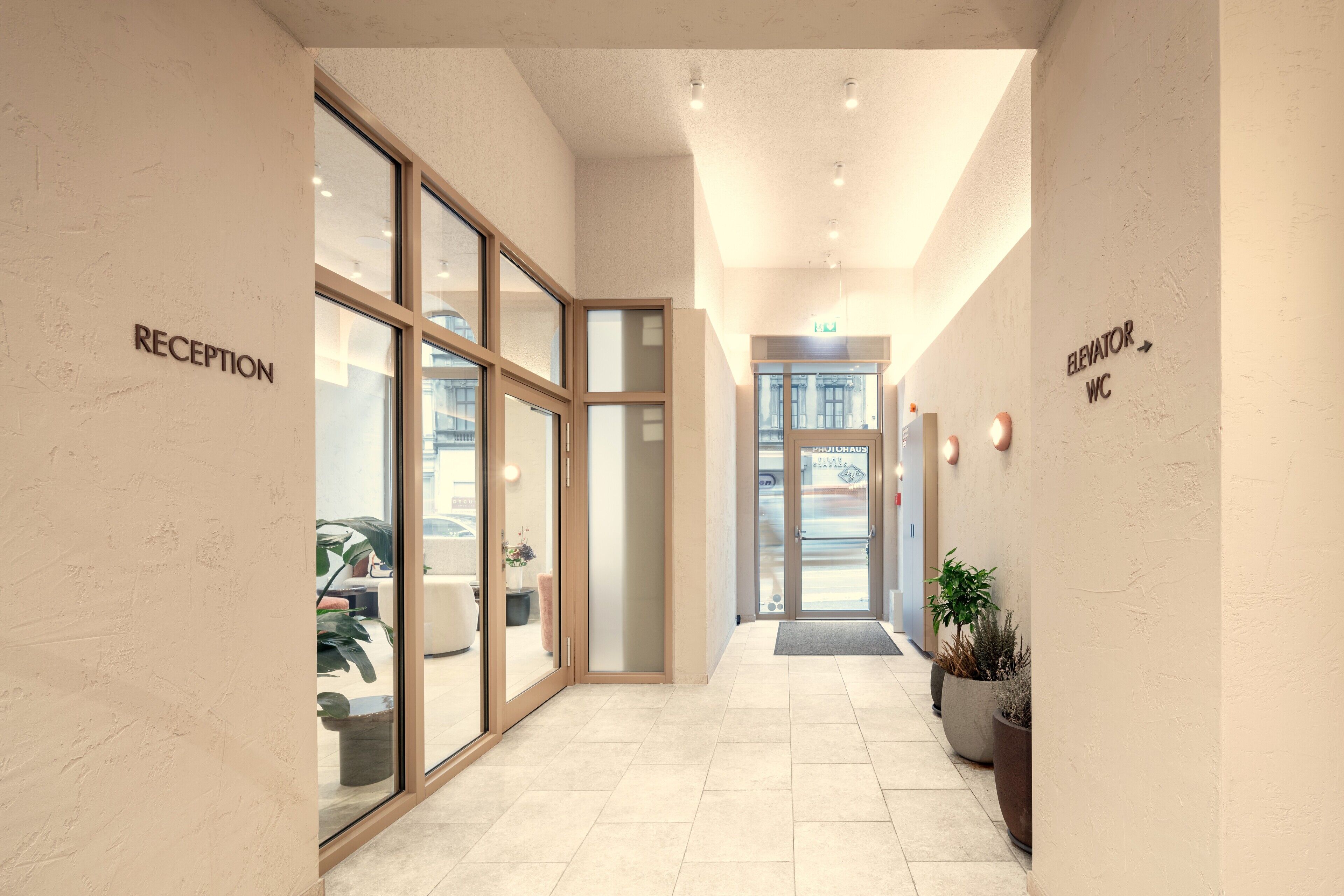 Uma área de recepção de hotel com entrada por porta de vidro, balcão de recepção e placa indicando a localização do elevador e sanitários.