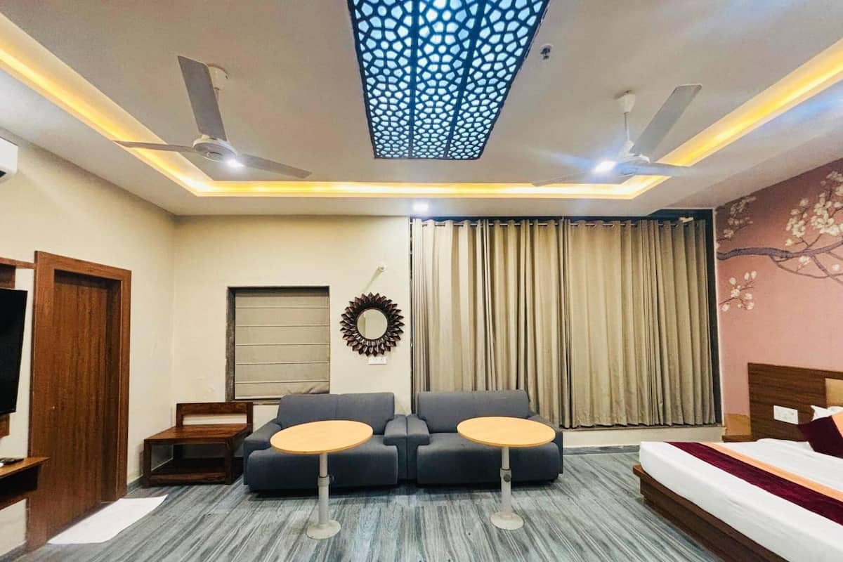 Quarto de hotel moderno com sofá, duas mesinhas, cama, televisão, ventoinha no teto e uma peça decorativa na parede.