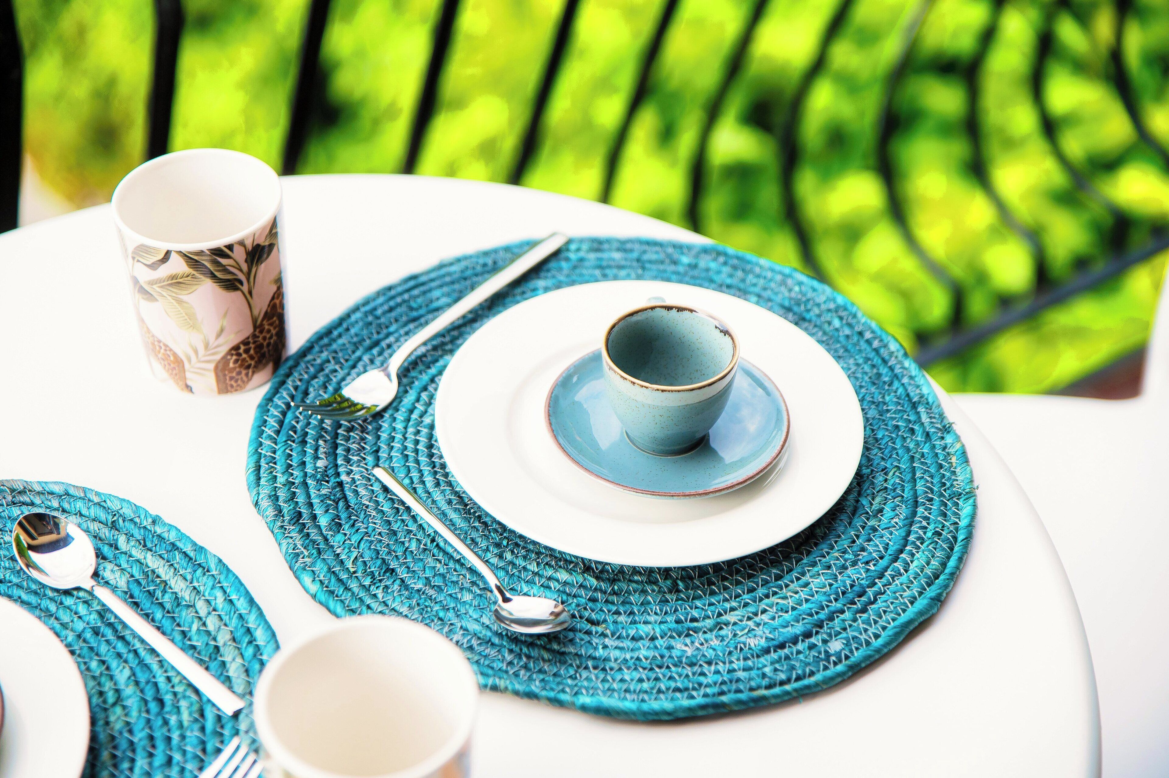 Mesa posta com um jogo de mesa azul tecido, um prato branco, uma xícara azul e uma colher de prata.