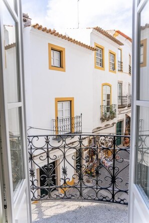 Vista de um edifício com paredes brancas e molduras de janelas amarelas, uma varanda com grades de metal preto ornamentadas e chão de pedra.