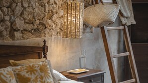 Quarto com parede de pedra, cama de madeira, mesa de cabeceira com abajur, cesto trançado e uma escada.