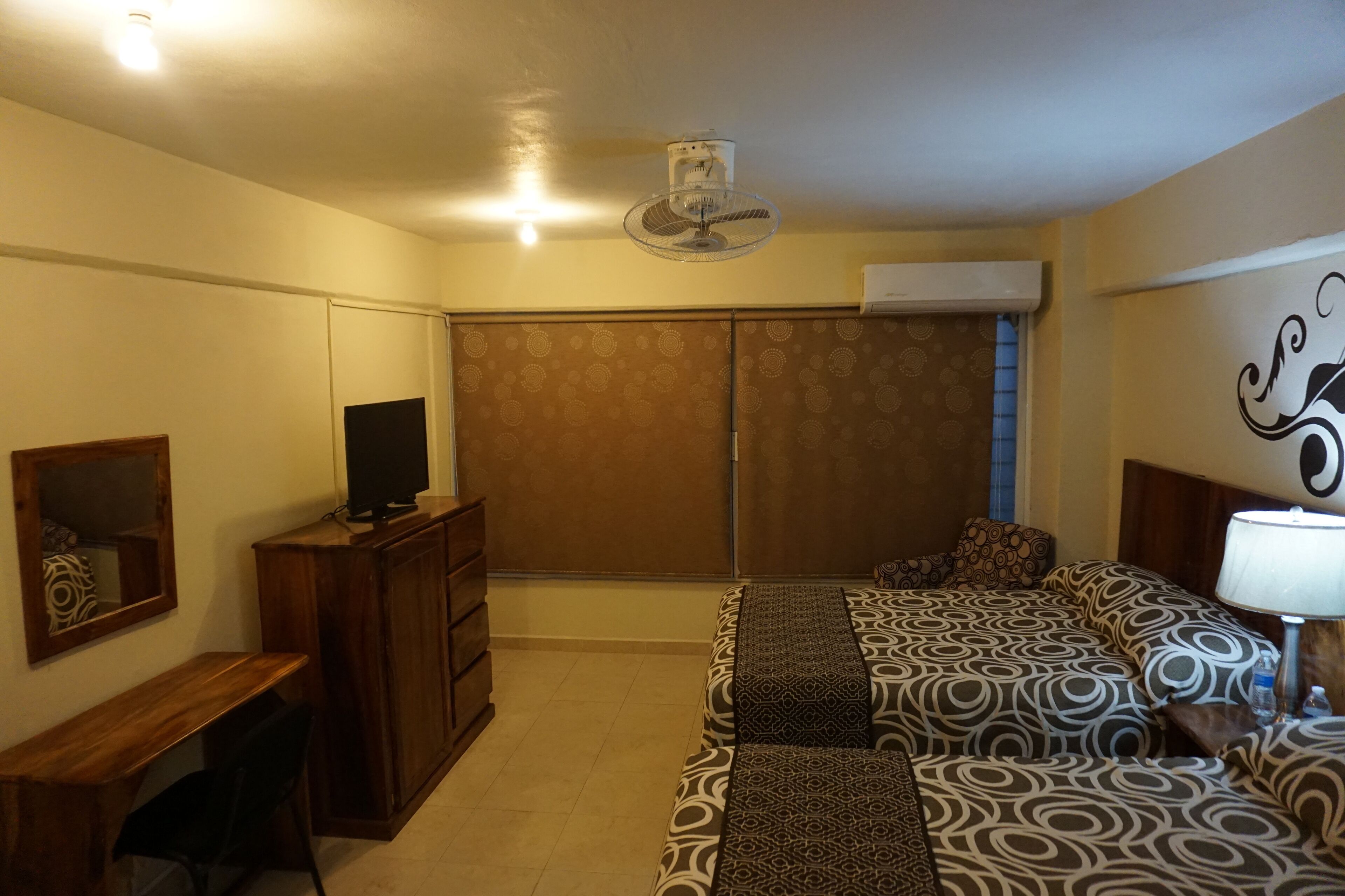 Quarto com cama, escrivaninha, televisão, guarda-roupa e ventoinha no teto.