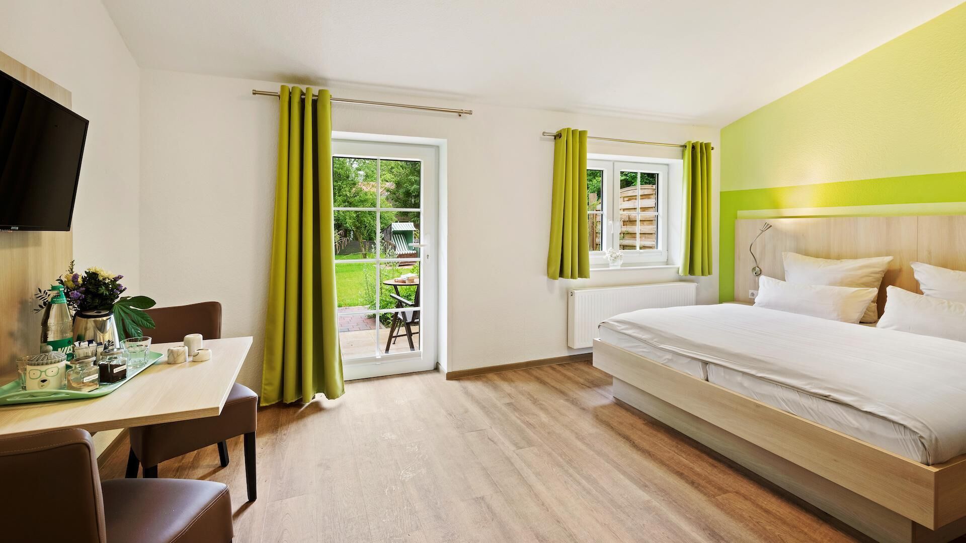 Quarto de hotel com cama, secretária com cadeira, televisão e janela com cortinas.