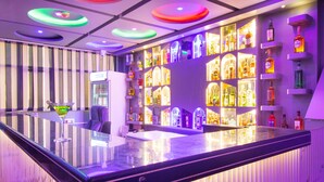 Uma área de bar com um balcão comprido, prateleiras com garrafas e uma geladeira.