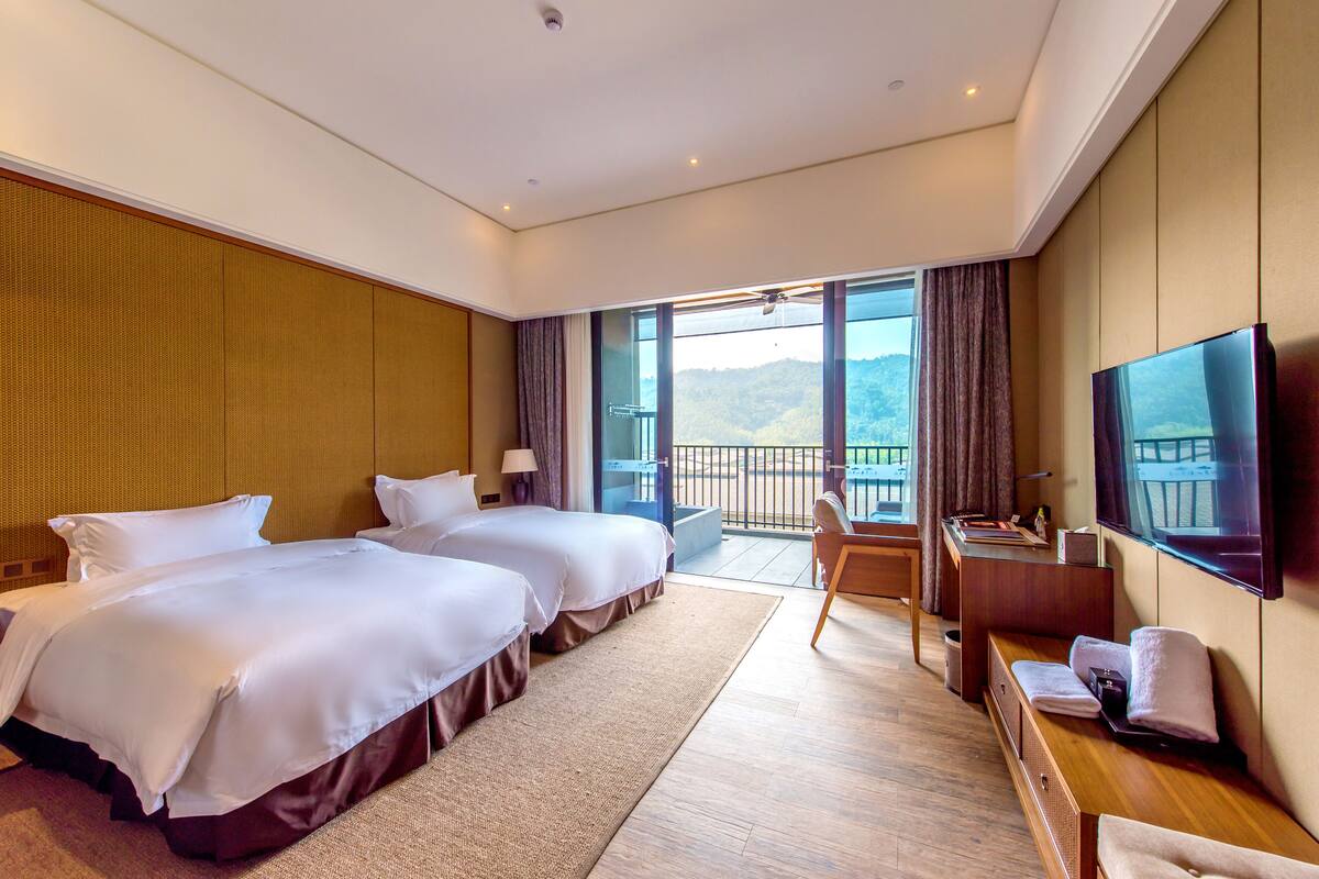 Quarto de hotel com duas camas, uma escrivaninha, televisão e varanda com vista para as montanhas.