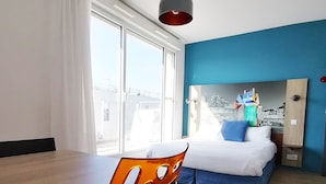 Quarto de hotel com uma janela grande, cama com roupa de cama azul, mesa de madeira e uma cadeira.