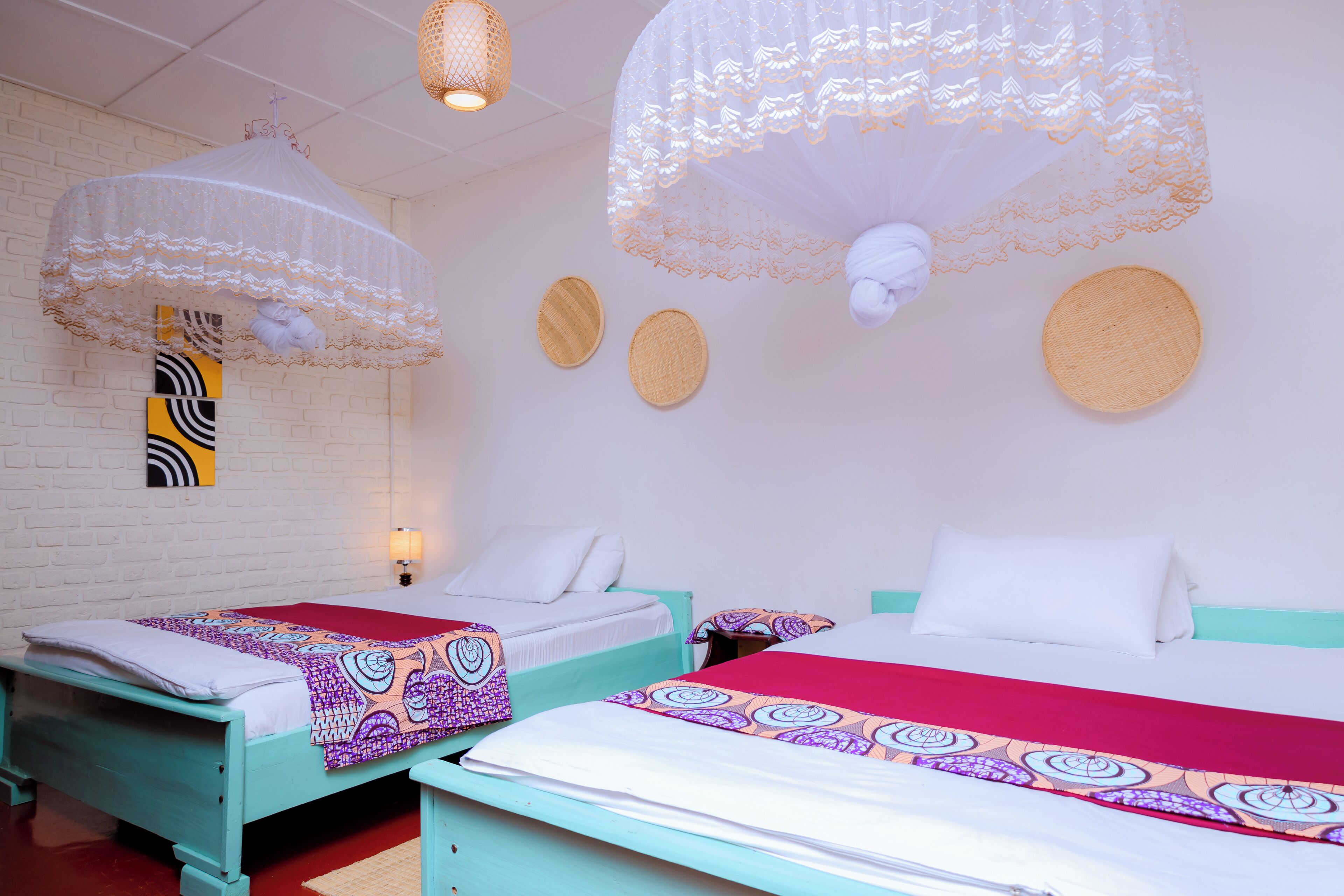Duas camas com roupa de cama branca, um edredom estampado em vermelho e roxo, e um travesseiro branco em cada cama. Há duas decorações de parede trançadas e uma parede branca com uma pintura abstrata em preto e branco.