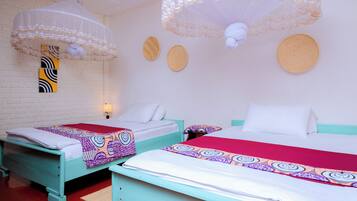 Duas camas com roupa de cama branca, um edredom estampado em vermelho e roxo, e um travesseiro branco em cada cama. Há duas decorações de parede trançadas e uma parede branca com uma pintura abstrata em preto e branco.