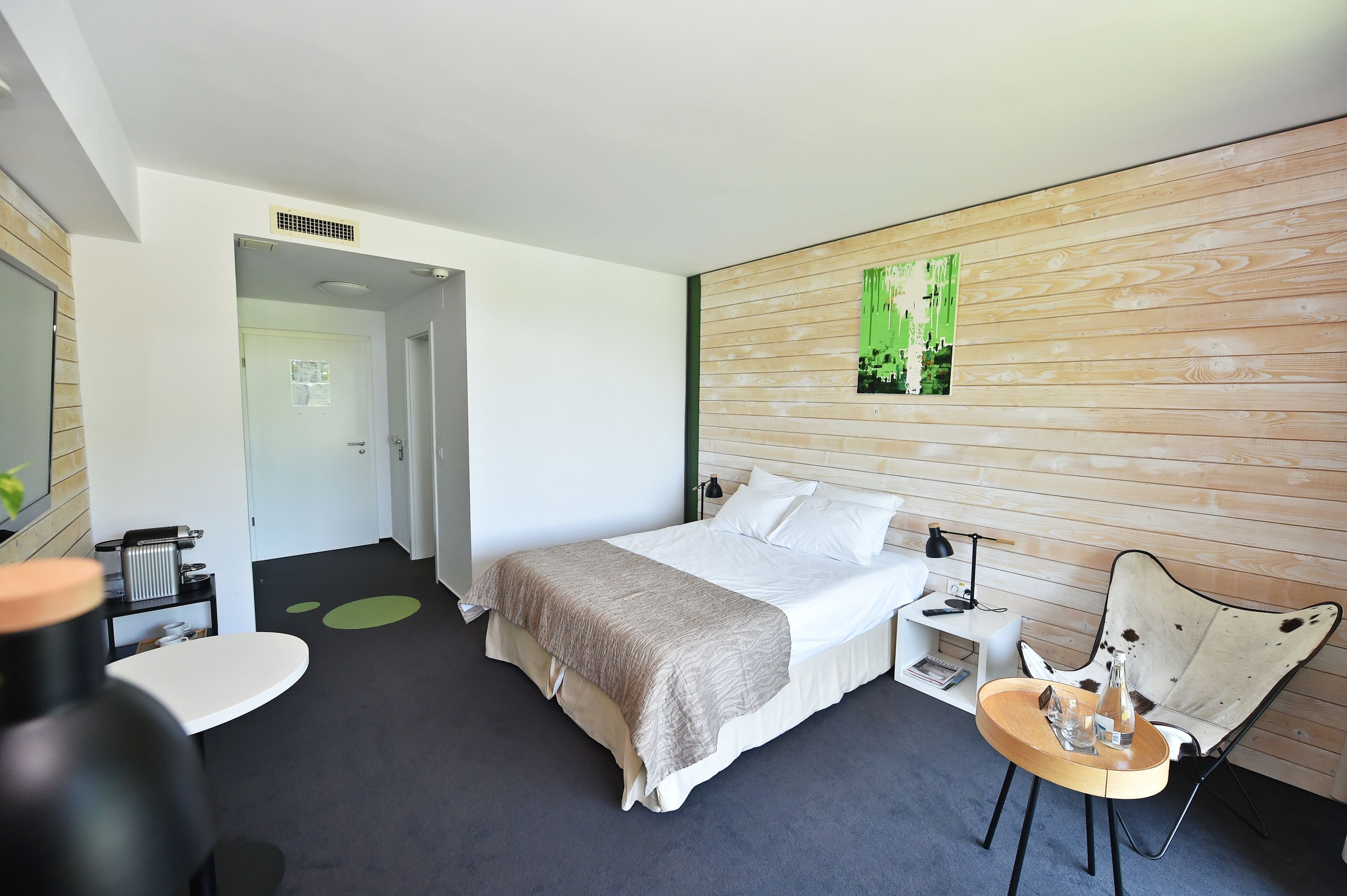 Quarto de hotel moderno com cama, mesa de cabeceira, cadeira e uma pequena mesa com garrafa e copos.