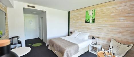 Quarto de hotel moderno com cama, mesa de cabeceira, cadeira e uma pequena mesa com garrafa e copos.