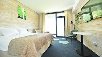 Quarto de hotel com uma cama grande, uma mesinha, uma cadeira e uma televisão fixada na parede.