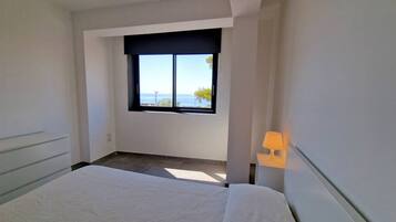 Quarto com uma janela grande, cama, mesa de cabeceira com abajur e vista para o mar.
