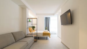 Quarto moderno e minimalista com sofá cinzento, cama com roupa de cama amarela, planta verde sobre uma estante preta e televisão de tela plana na parede.