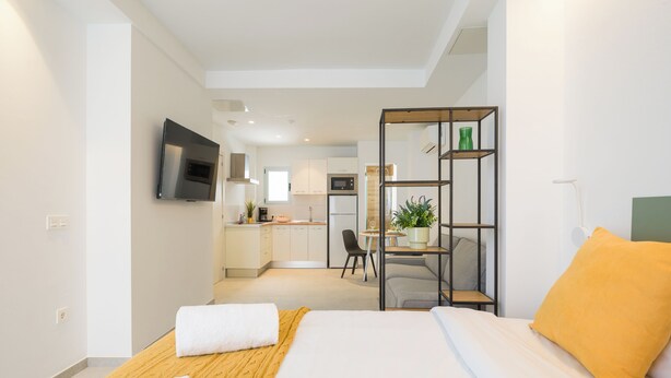 Quarto de hotel moderno com cama, sofá, televisão na parede, área de cozinha e mesa de jantar com cadeiras.