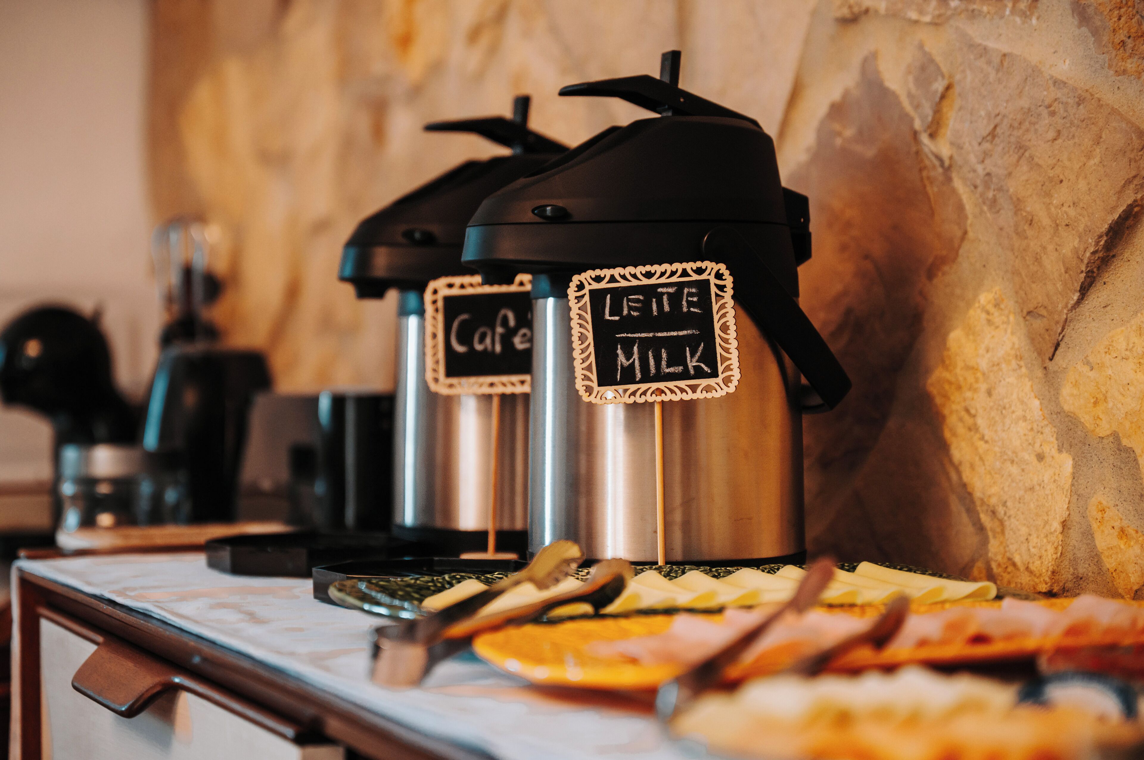 Buffet com dispensadores de café e leite identificados, variedade de alimentos e parede de pedra ao fundo.