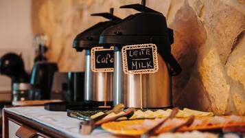 Buffet com dispensadores de café e leite identificados, variedade de alimentos e parede de pedra ao fundo.