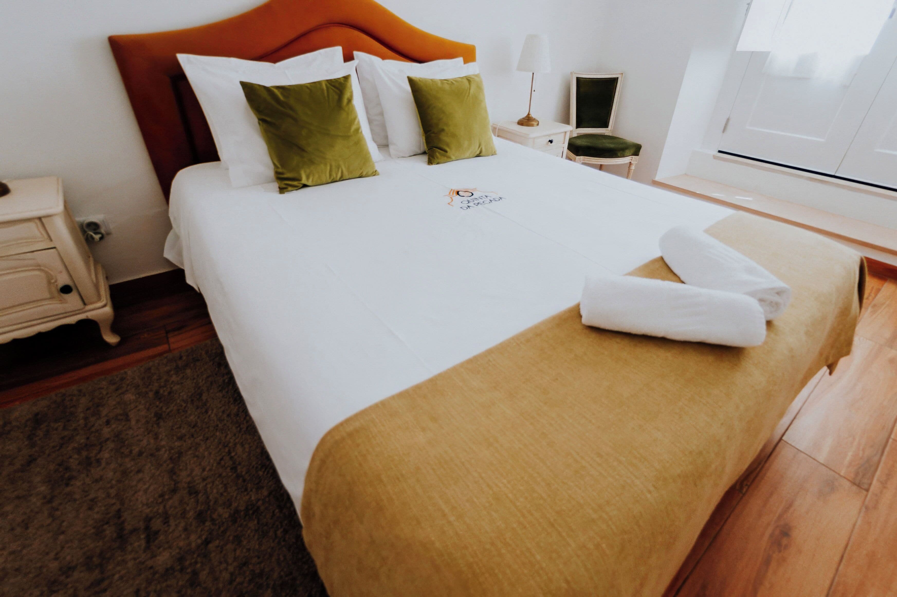 Cama bem arrumada com travesseiros brancos e verdes, um cobertor marrom e uma toalha dobrada ao lado.