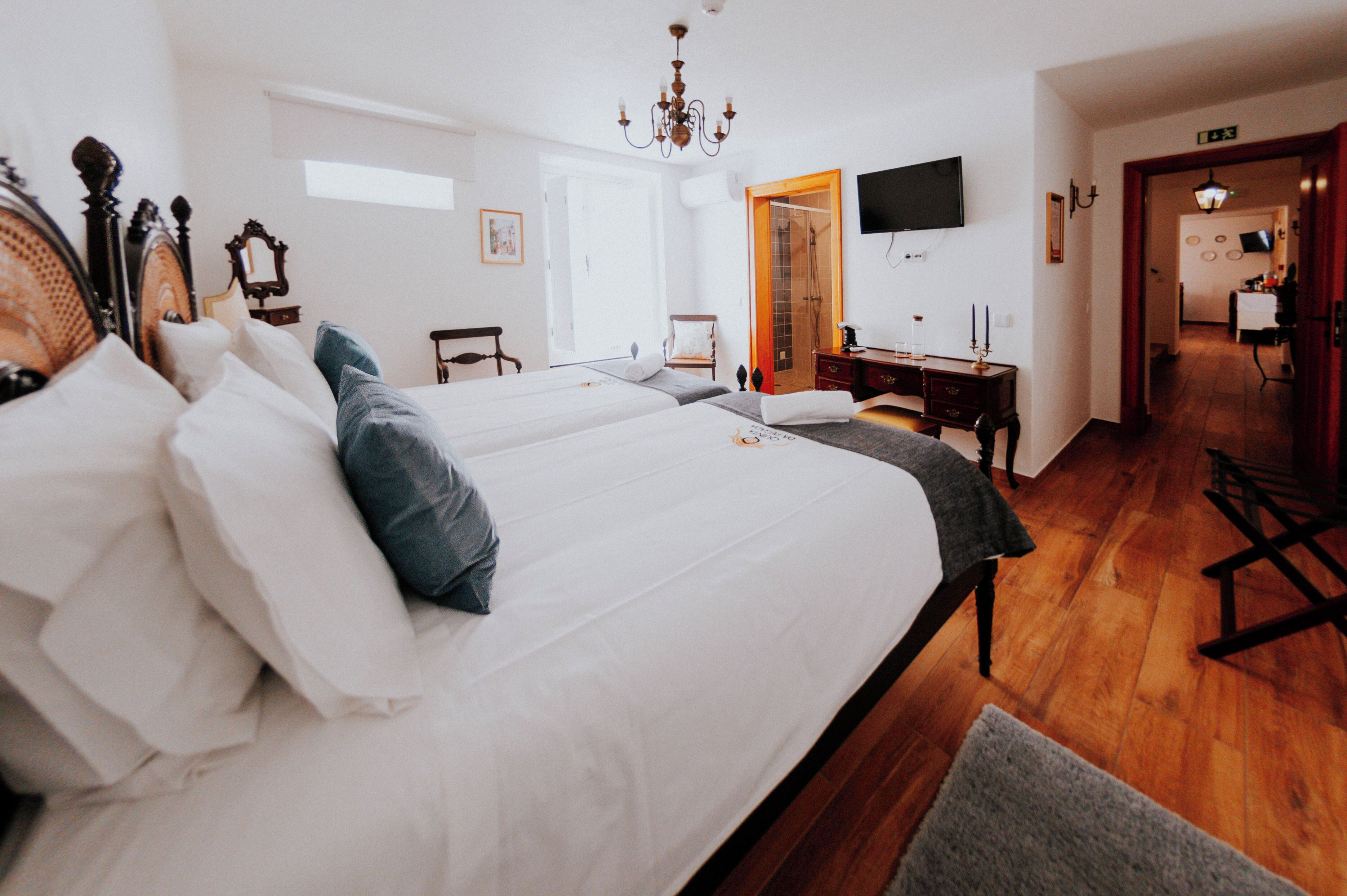 Quarto de hotel com uma cama grande, escrivaninha, cadeira, televisão e um candelabro.