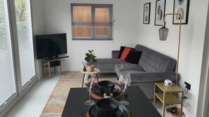 Uma sala moderna com um sofá cinzento, uma TV de tela plana, uma mesa de jantar posta para dois e uma grande janela com persianas.