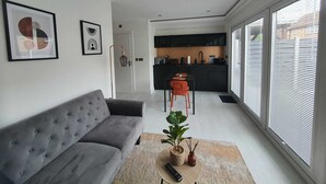 Uma sala de estar moderna com um sofá cinzento, uma mesa de centro com uma planta e uma cozinha ao fundo.