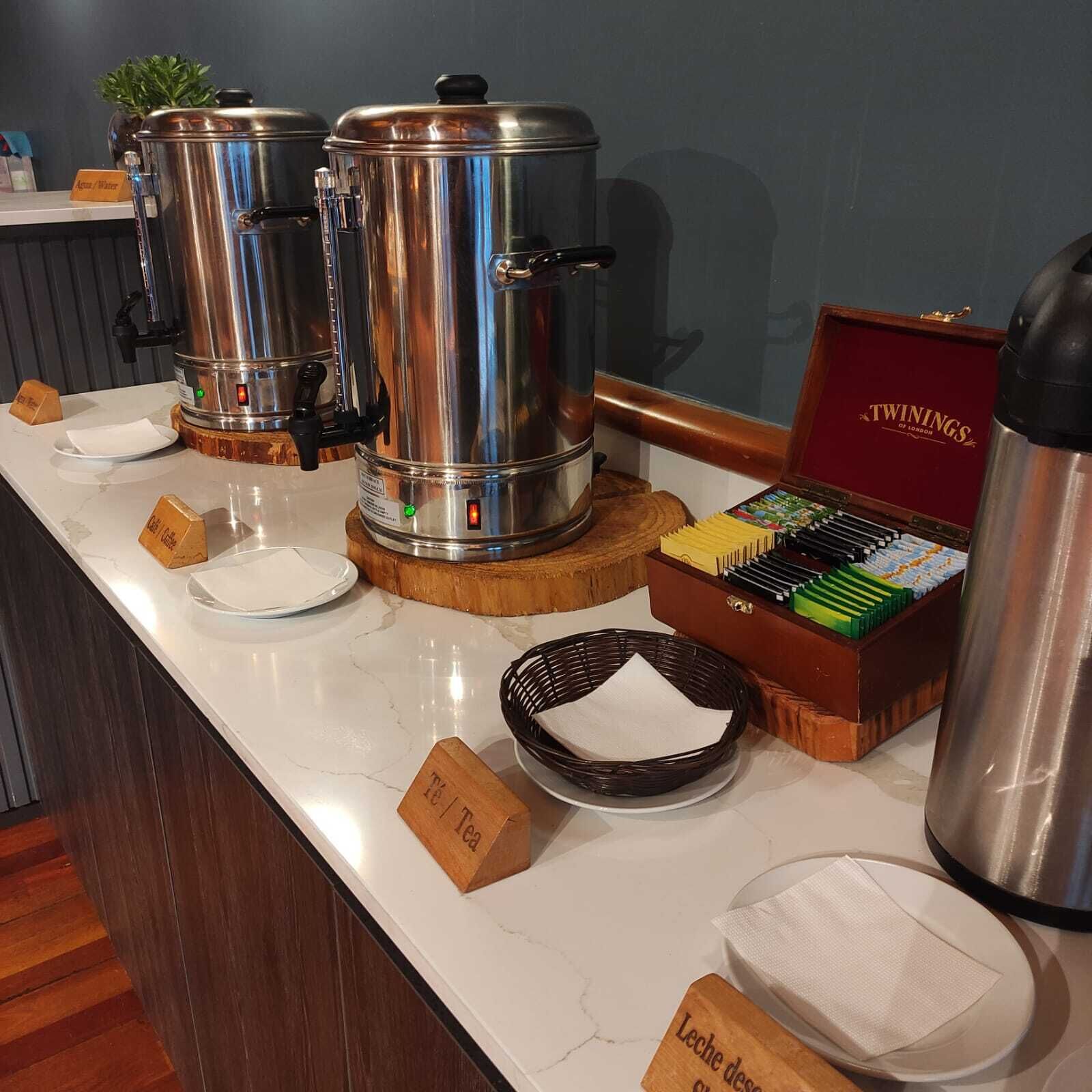 Disposição de buffet com várias máquinas de café em aço inoxidável, dispensadores de chá e café, e uma variedade de lanches embalados.