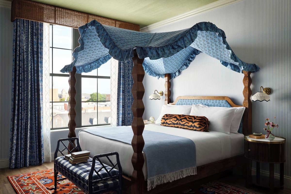 Uma cama com dossel e cortinas, vista para a cidade através de grandes janelas, uma mesa de cabeceira com um vaso de flores e um tapete estampado.