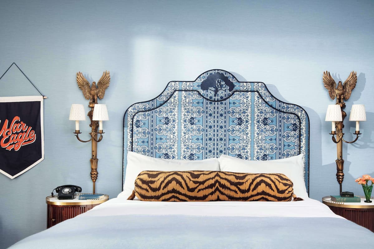 Quarto com cabeceira estampada, duas arandelas de parede, um quadro, um telefone antigo e uma pequena mesa com um vaso de flores.