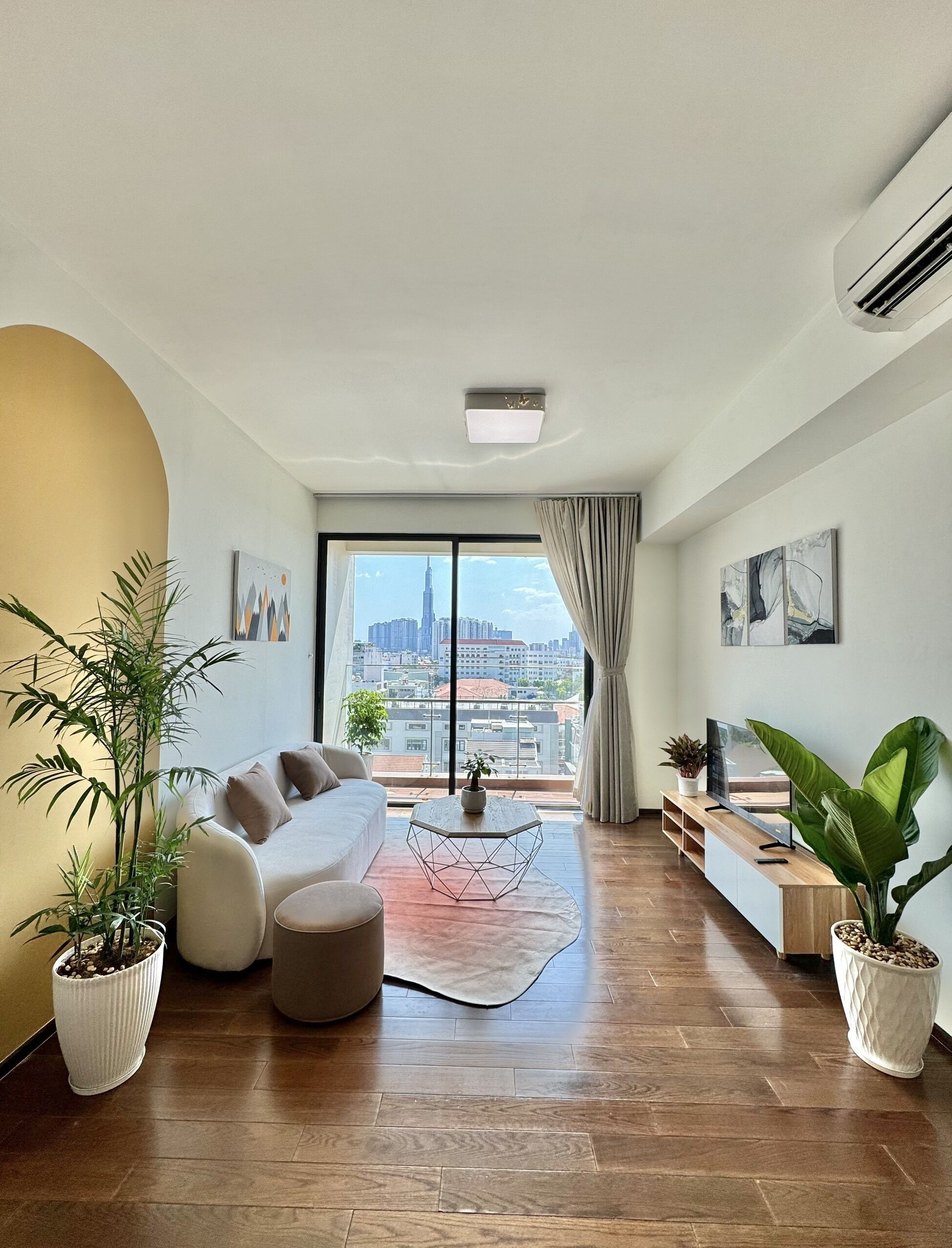 Uma sala de estar moderna com sofá, mesa de centro e plantas em vasos. Uma janela ampla oferece vista para a cidade.