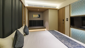 Quarto de hotel moderno com uma cama grande, área de estar com um banco acolchoado, televisão e um separador decorativo.