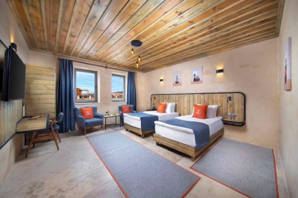 Quarto de hotel com vigas de madeira no teto, duas camas com roupa de cama azul e laranja, uma mesa pequena com cadeiras e uma janela grande.