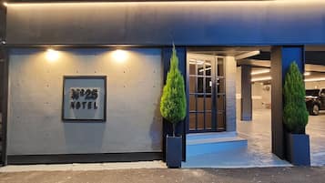 Entrada de hotel com uma placa indicando "No.25 HOTEL", duas plantas em vasos e uma garagem de estacionamento ao fundo.