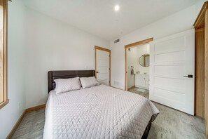 Quarto com uma cama grande, cabeceira de madeira, paredes brancas e piso de madeira. Duas portas brancas e um banheiro são visíveis ao fundo.