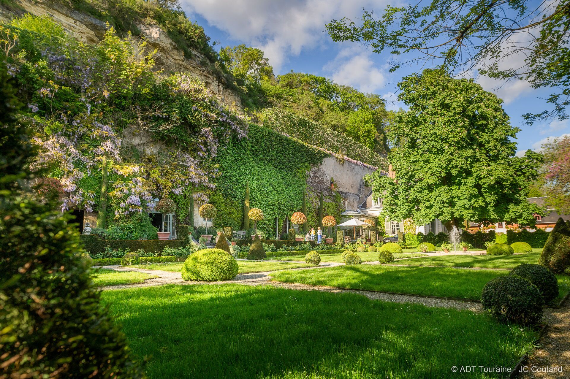 Um jardim exuberante com sebes bem aparadas, diversas árvores e um edifício de fachada em pedra parcialmente coberto por hera.