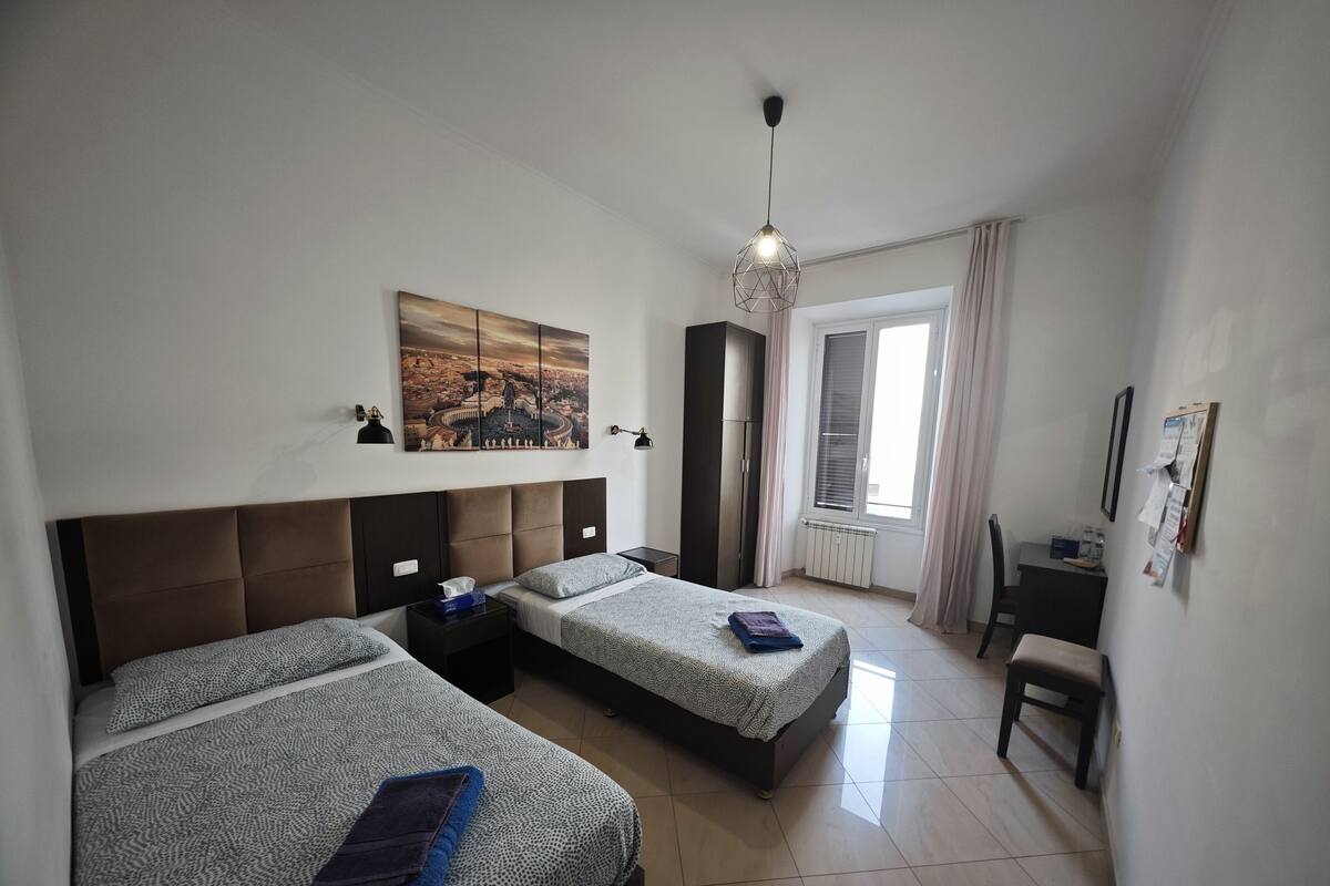 Quarto de hotel com duas camas, um quadro grande, um radiador, uma escrivaninha e uma cadeira.