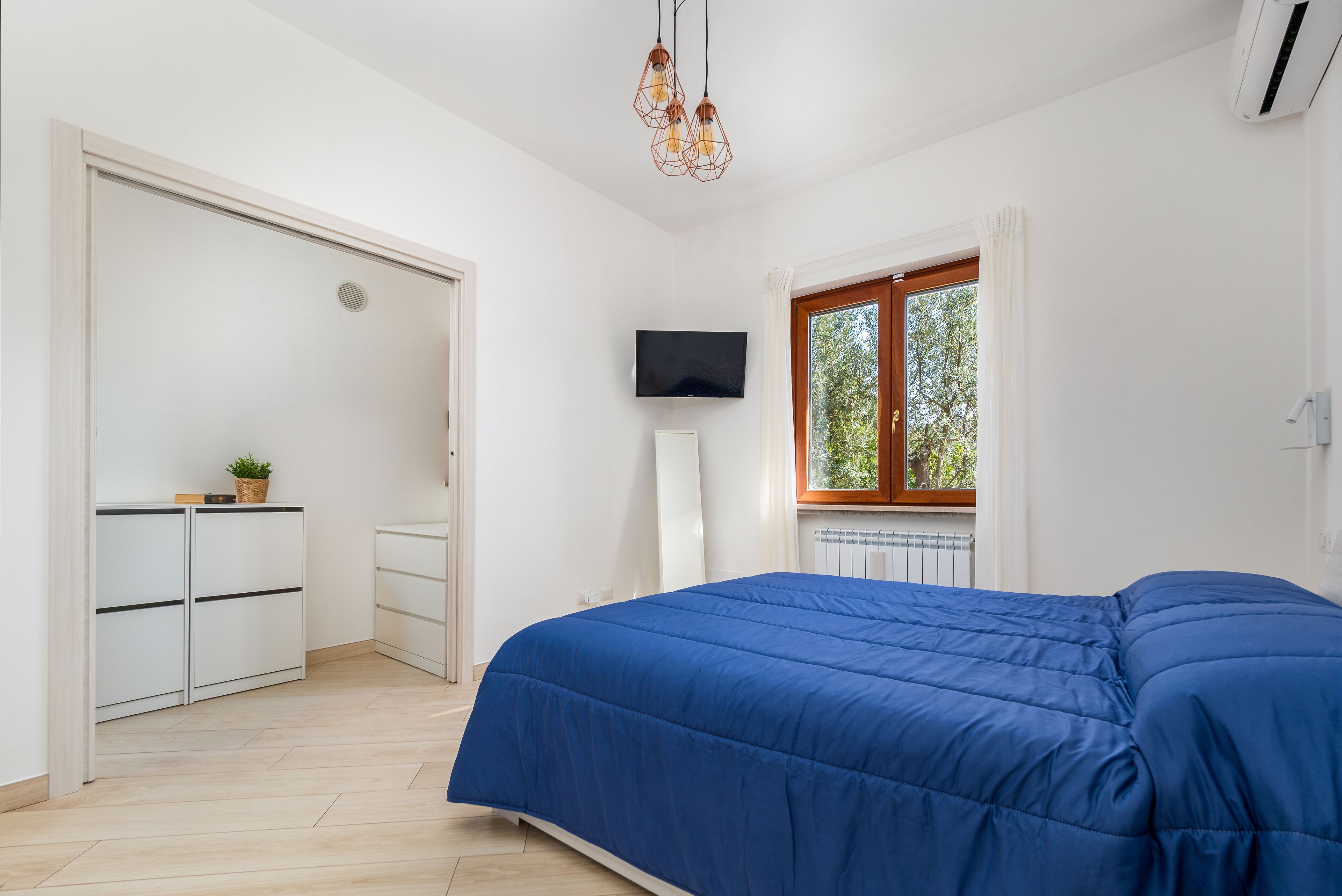Quarto com uma cama grande, televisão na parede e janela com vista para o verde.