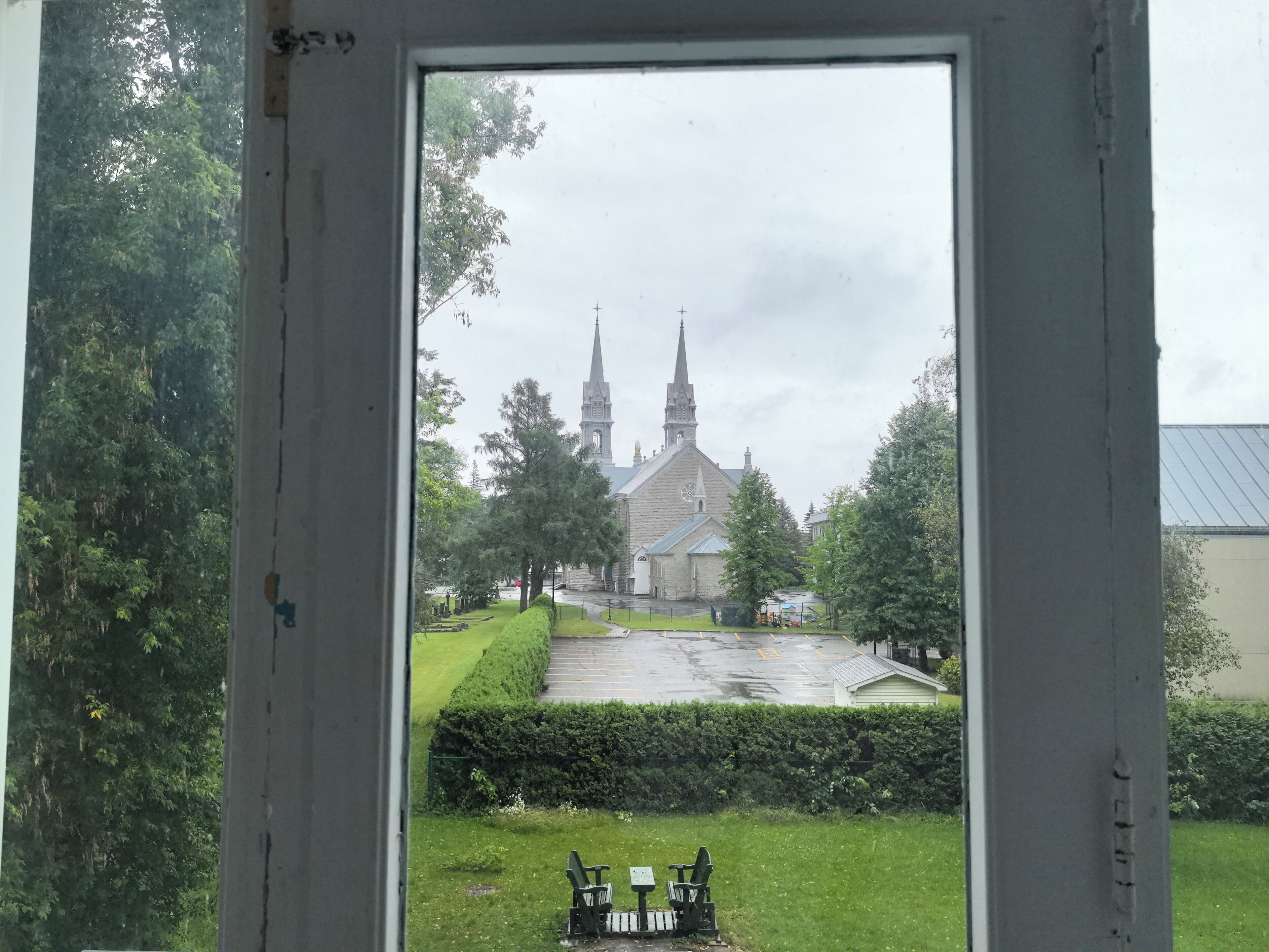 Visão pela janela de uma igreja com duas torres, um gramado verde e um banco.
