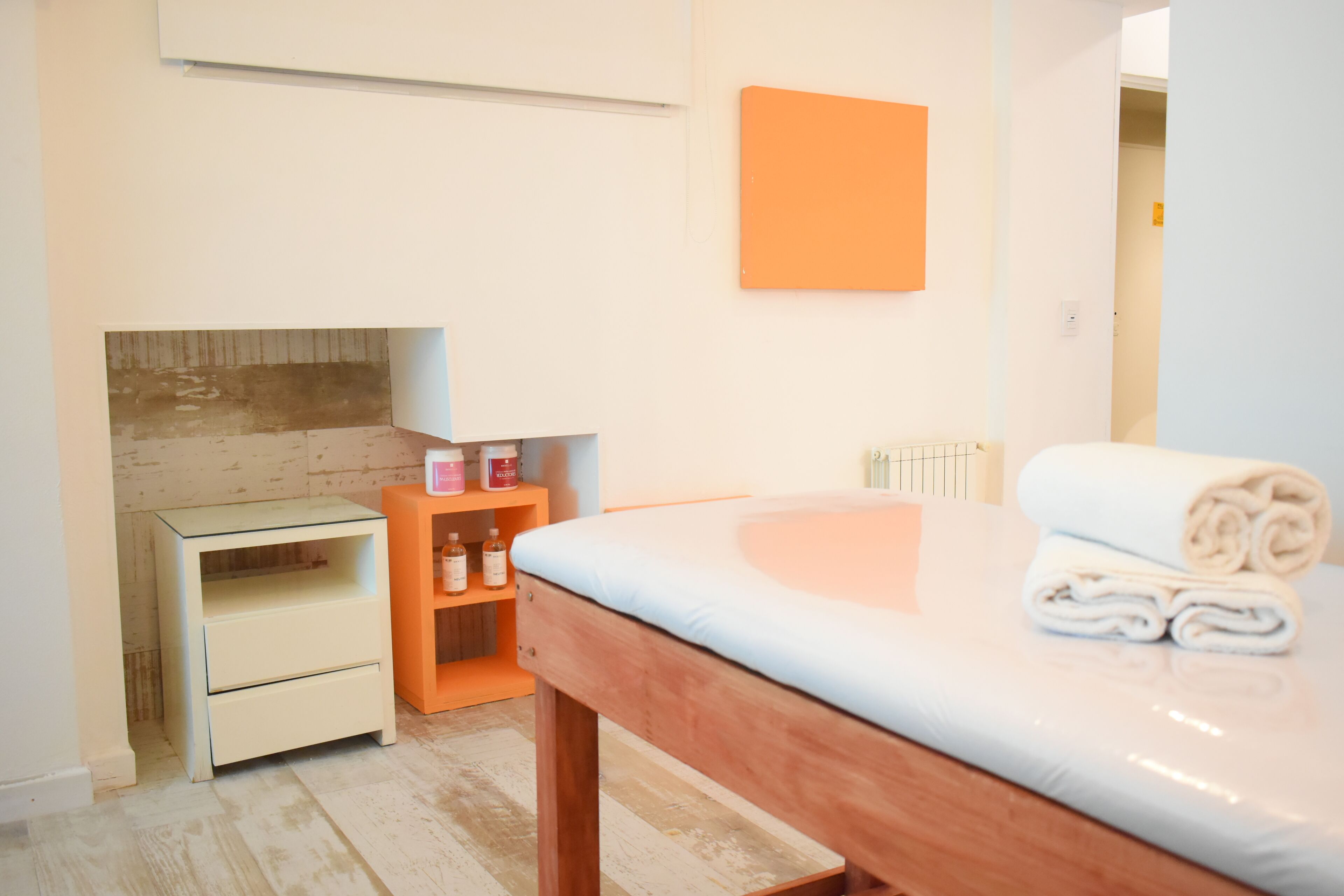 Quarto de hotel com cama de madeira, mesa de cabeceira, prateleira pequena com artigos de higiene e painel laranja fixado na parede.