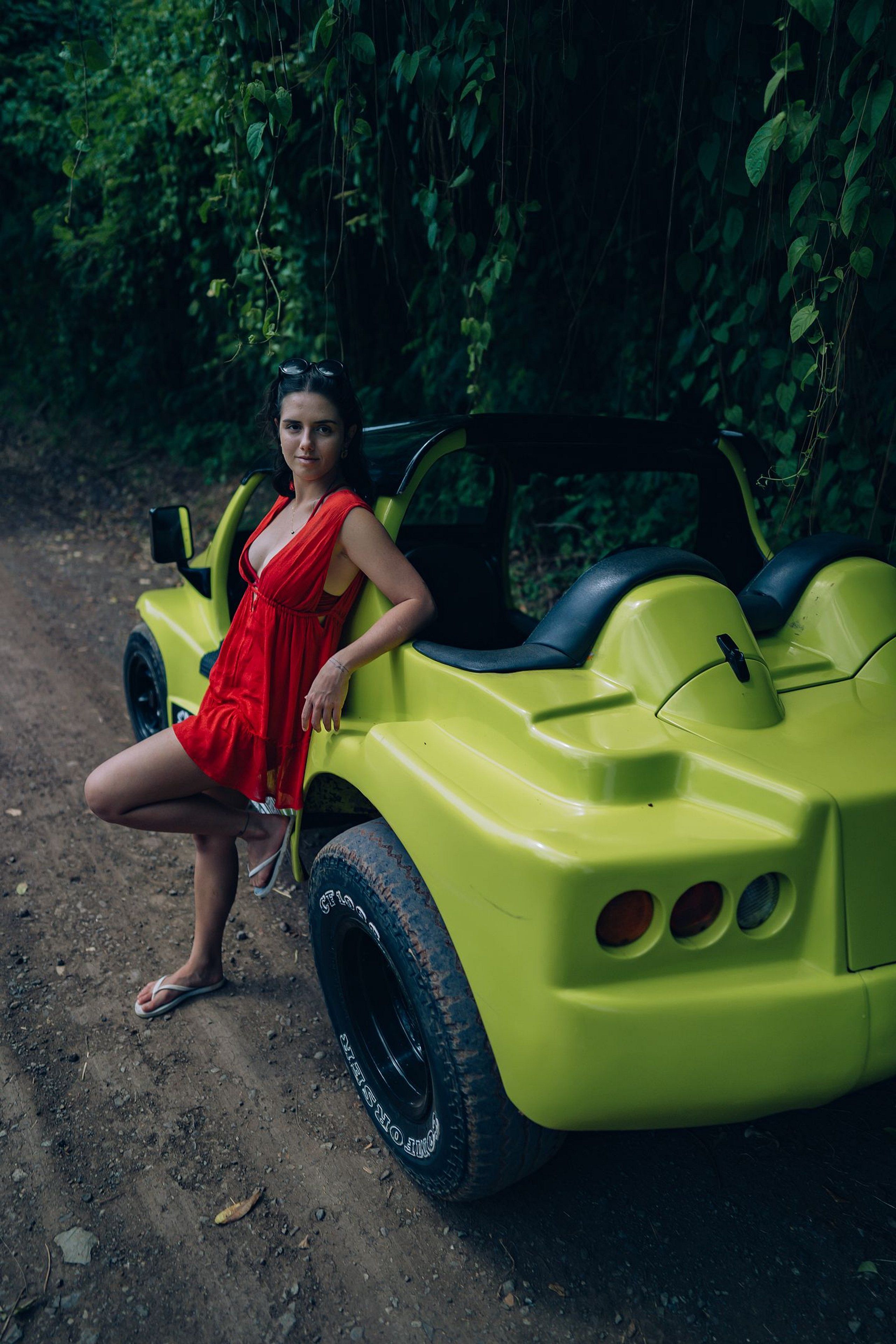 Uma pessoa de vestido vermelho apoiada em um veículo amarelo vibrante numa estrada de terra, cercada por vegetação densa.