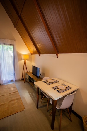 Um quarto aconchegante com teto de madeira, uma TV sobre suporte, uma mesa de jantar com cadeiras e um abajur.