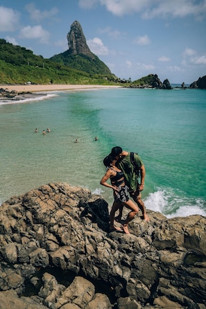 Uma linha costeira rochosa com águas turquesa cristalinas, uma montanha ao longe e uma praia ao fundo.