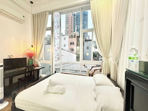 Quarto de hotel com cama, escrivaninha com televisão, janela com cortinas e vista para prédios.