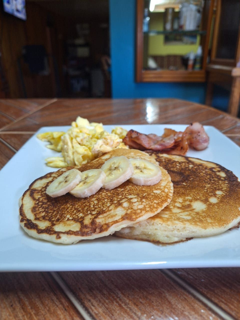 Prato de café da manhã com panquecas, ovos mexidos, bacon e fatias de banana sobre um prato branco.