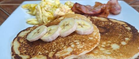Prato de café da manhã com panquecas, ovos mexidos, bacon e fatias de banana sobre um prato branco.
