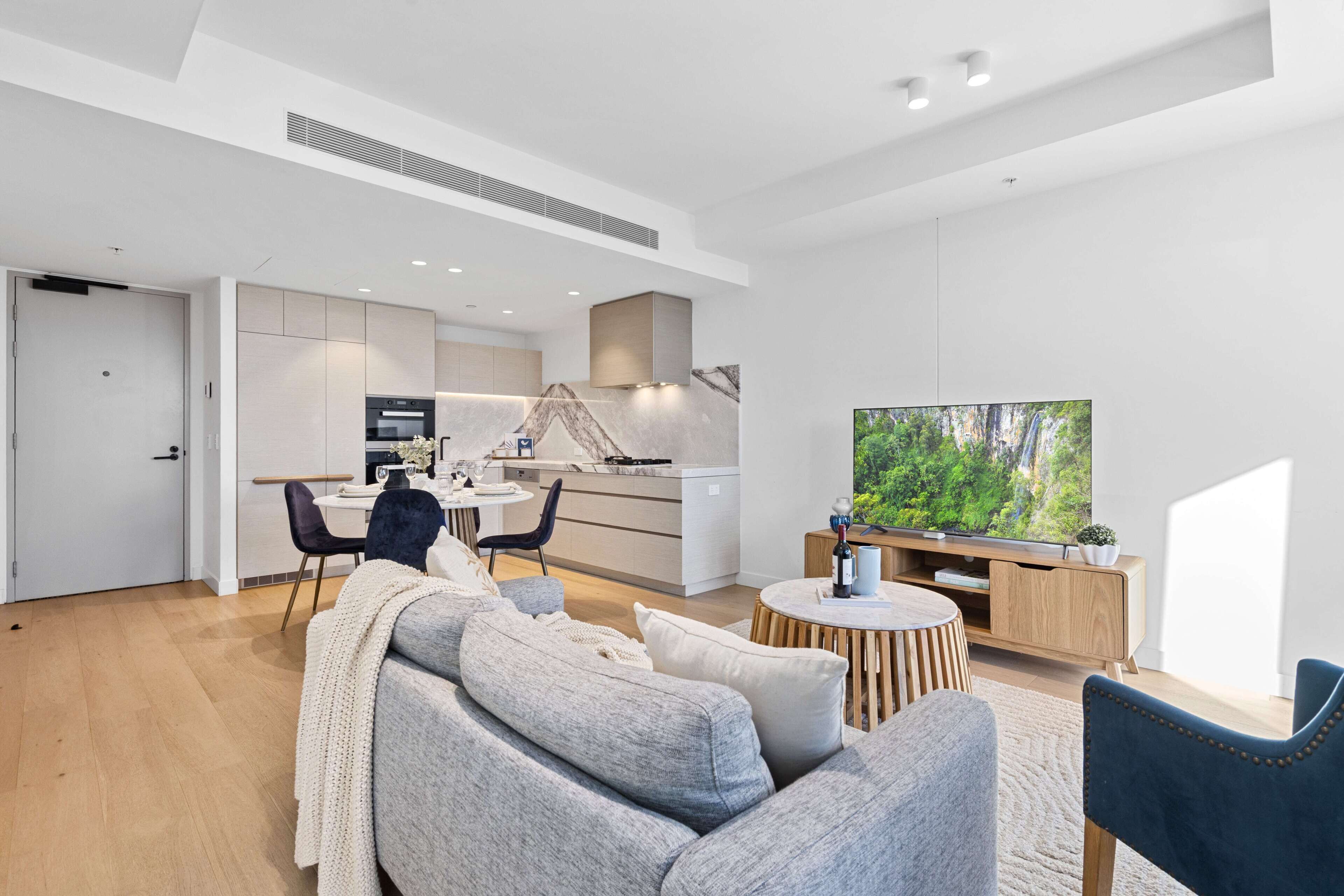 Uma sala de estar moderna com sofá, mesa de centro e televisão. Cozinha visível ao fundo, com armários e bancada.