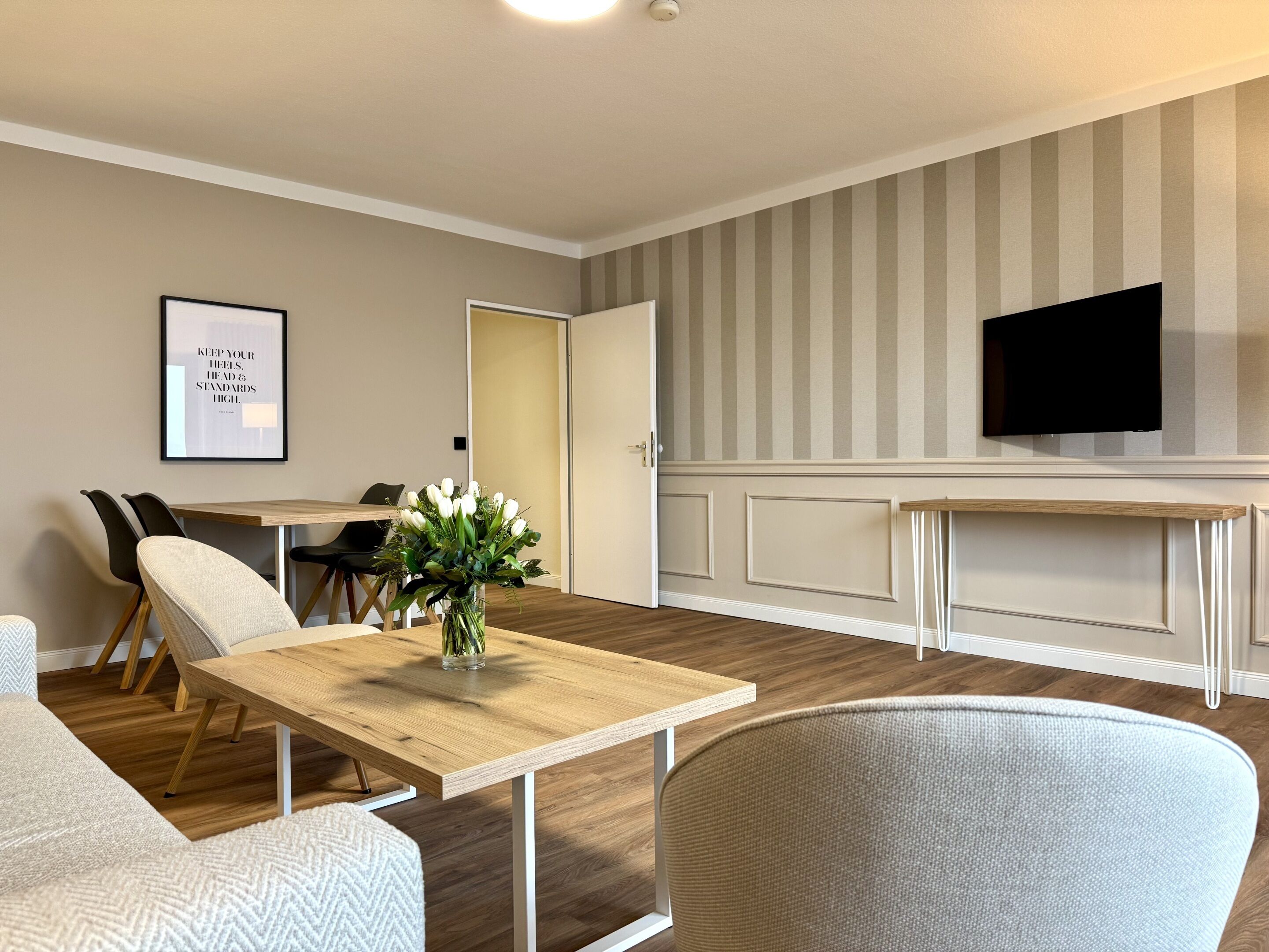 Uma sala de estar moderna com uma mesa de jantar de madeira, duas cadeiras, um sofá, uma TV fixada na parede e uma frase emoldurada na parede.