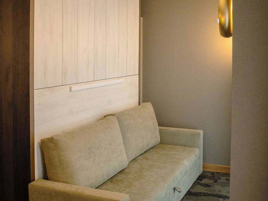 Um sofá em tons neutros com cabeceira de madeira, num quarto com carpete estampado e luminária fixada na parede.