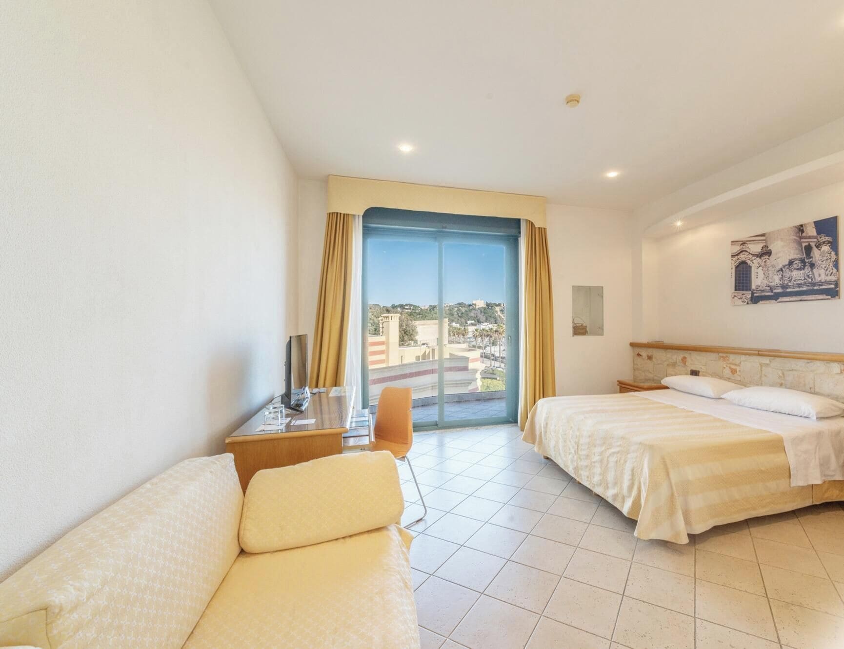 Quarto de hotel com cama, sofá, escrivaninha iluminada por uma luminária, janela com cortinas e um quadro na parede.